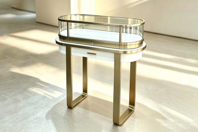 China Custom Luxury Oval Curved Glass Jewelry Display Table with Brushed Gold Metal Leg and Lockable Storage Drawer for sale